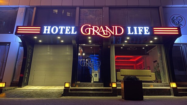 Exterior do HOTEL GRAND LIFE, com letreiros iluminados, porta de garagem fechada e entrada bem iluminada.