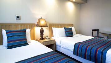 Quarto de hotel com duas camas, cada uma com um cobertor listrado e travesseiros brancos. Há uma mesa de cabeceira com abajur e telefone.