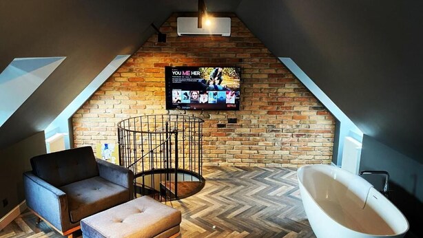 Quarto moderno estilo loft com piso de madeira em padrão herringbone, uma banheira branca grande, uma gaiola de metal preta, um sofá cinza e uma TV montada em parede de tijolos exibindo 'YOU ME HER'.
