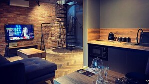 Uma sala de estar moderna com parede de tijolos, escada, TV de tela plana, mesa de centro de madeira e área de cozinha com micro-ondas e cafeteira.