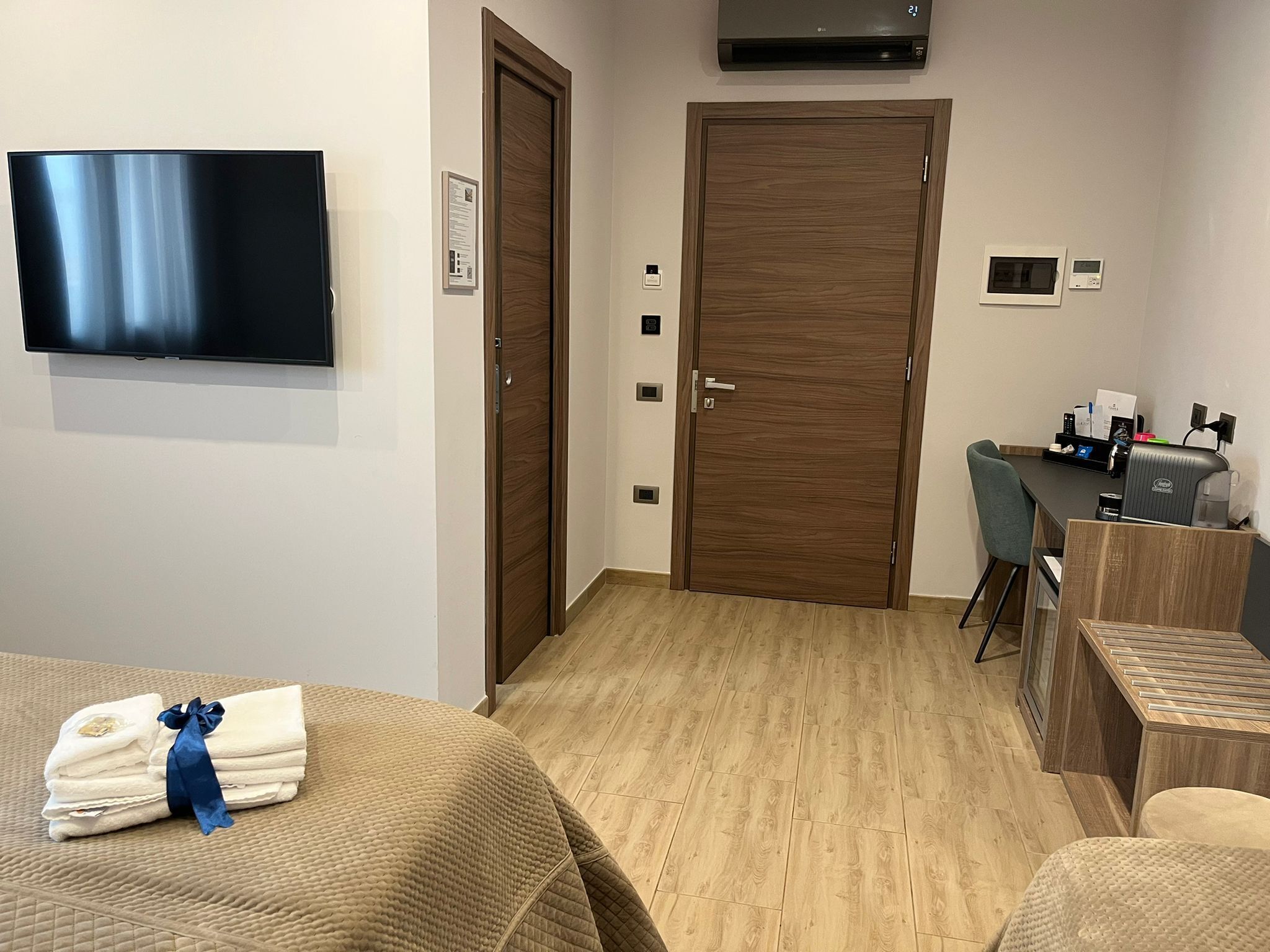 Quarto de hotel com uma cama, uma escrivaninha com cadeira, uma televisão e uma porta de madeira.