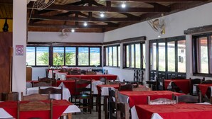 Restaurante com vigas de madeira e grandes janelas oferecendo vista para o exterior. Mesas dispostas com toalhas vermelhas e guardanapos brancos.
