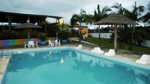 Uma piscina com cadeiras de descanso brancas e um guarda-sol de palha.
