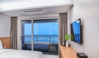 Quarto de hotel com cama, escrivaninha, televisão e porta de vidro que dá para uma varanda com vista para o oceano.