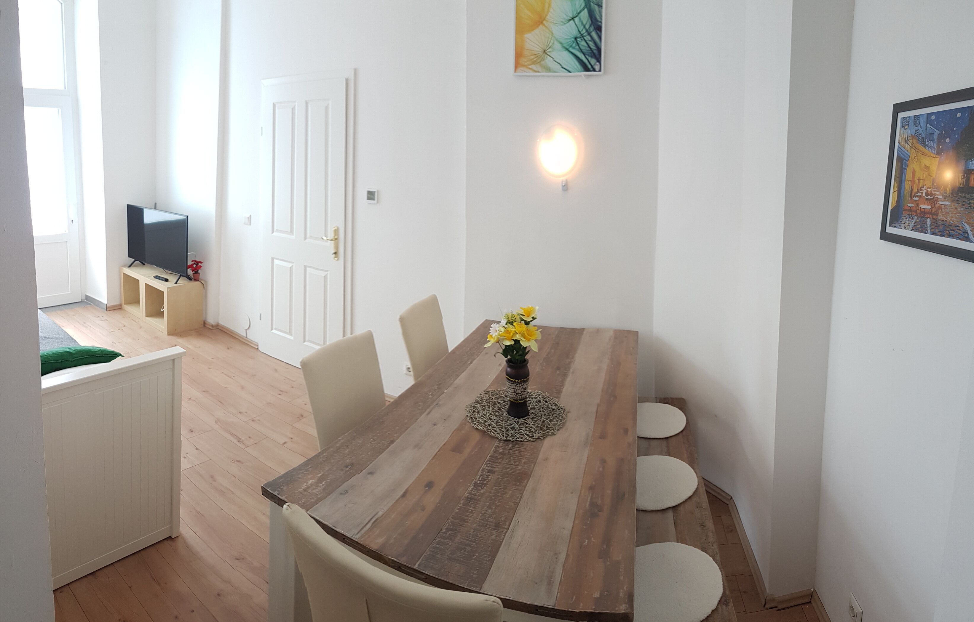 Uma sala de jantar com mesa de madeira, cadeiras brancas, um vaso com flores amarelas e um quadro na parede.