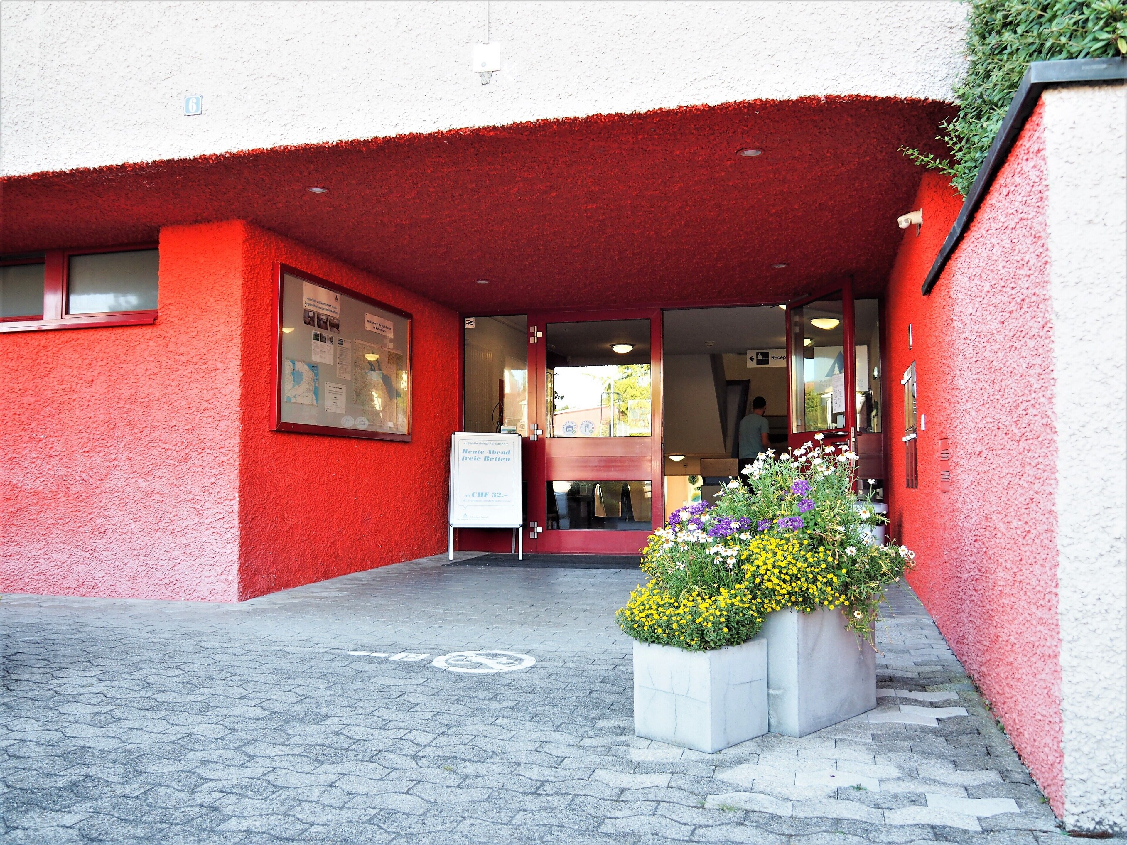 Entrada de um edifício vermelho com rampa acessível para cadeiras de rodas, balcão de recepção e vasos de flores.