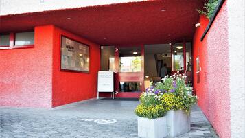 Entrada de um edifício vermelho com rampa acessível para cadeiras de rodas, balcão de recepção e vasos de flores.