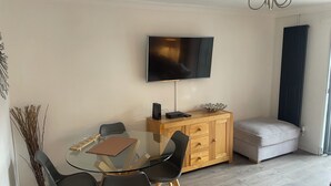 Uma sala de estar moderna com uma mesa de jantar de vidro, cadeiras pretas, um armário de madeira, uma televisão de tela plana e uma janela com cortinas.