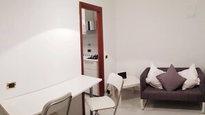 Um quarto pequeno com uma escrivaninha branca, uma cadeira, um sofá com almofadas e uma porta que dá para uma cozinha compacta.