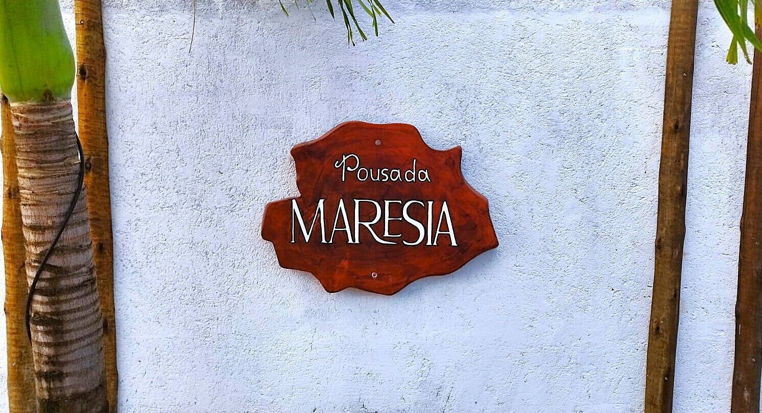 Placa de madeira com a inscrição 'Pousada da MARESIA' fixada em uma parede branca.