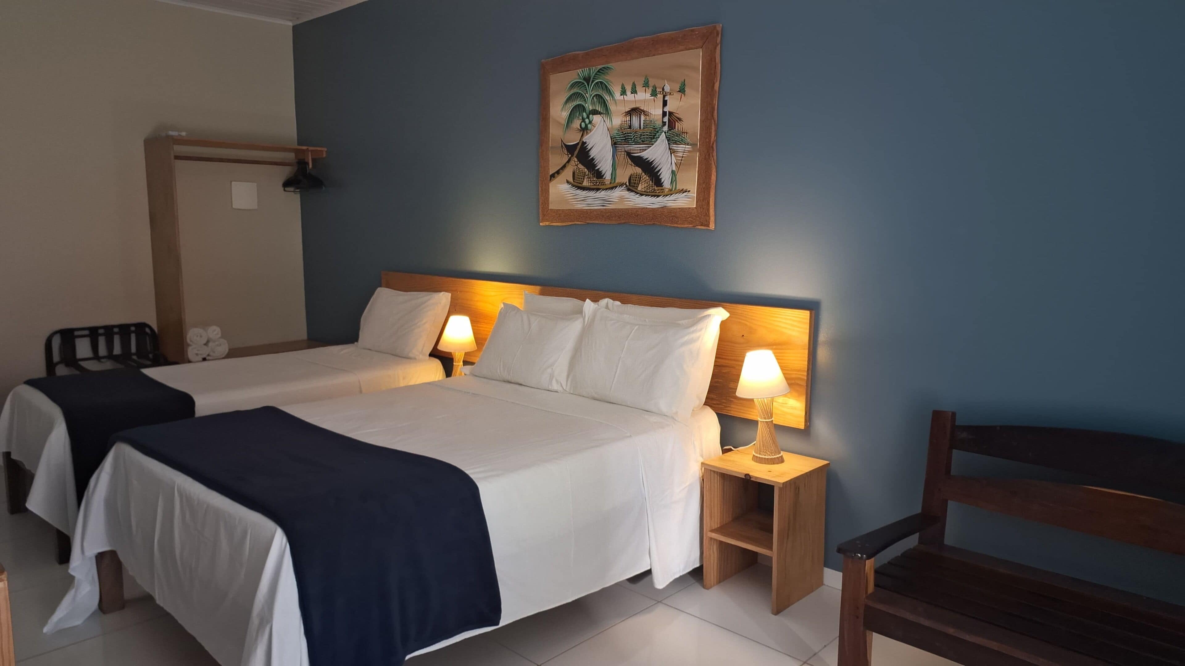 Quarto de hotel com duas camas, cabeceira de madeira, abajures, uma cadeira de madeira e uma pintura de paisagem tropical.
