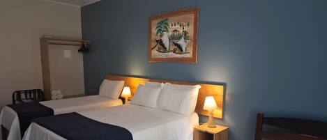 Quarto de hotel com duas camas, cabeceira de madeira, abajures, uma cadeira de madeira e uma pintura de paisagem tropical.