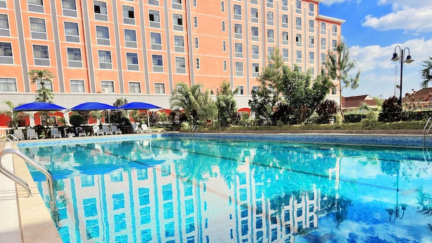 Piscina com água azul cristalina, rodeada por espreguiçadeiras e guarda-sóis, com um edifício de hotel ao fundo.