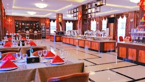 Restaurante com várias mesas de jantar dispostas com guardanapos e copos vermelhos, área de bar e balcão de buffet.