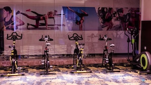 Uma academia com bicicletas ergométricas, equipamentos de exercício, paredes com espelhos e carpete estampado.
