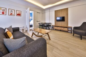 Uma sala de estar moderna com sofá, mesa de centro e televisão. Apresenta um painel de parede de madeira e uma mesa de jantar com cadeiras.