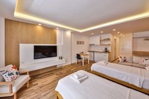 Quarto de hotel moderno com televisão de tela plana, duas camas, sofá, área de refeições e cozinha compacta.
