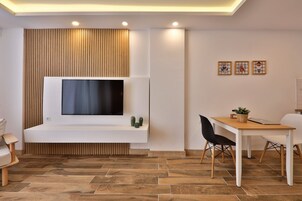 Uma sala de estar moderna com uma televisão de tela plana fixada em um console branco, uma mesa de jantar com cadeiras e piso de madeira.