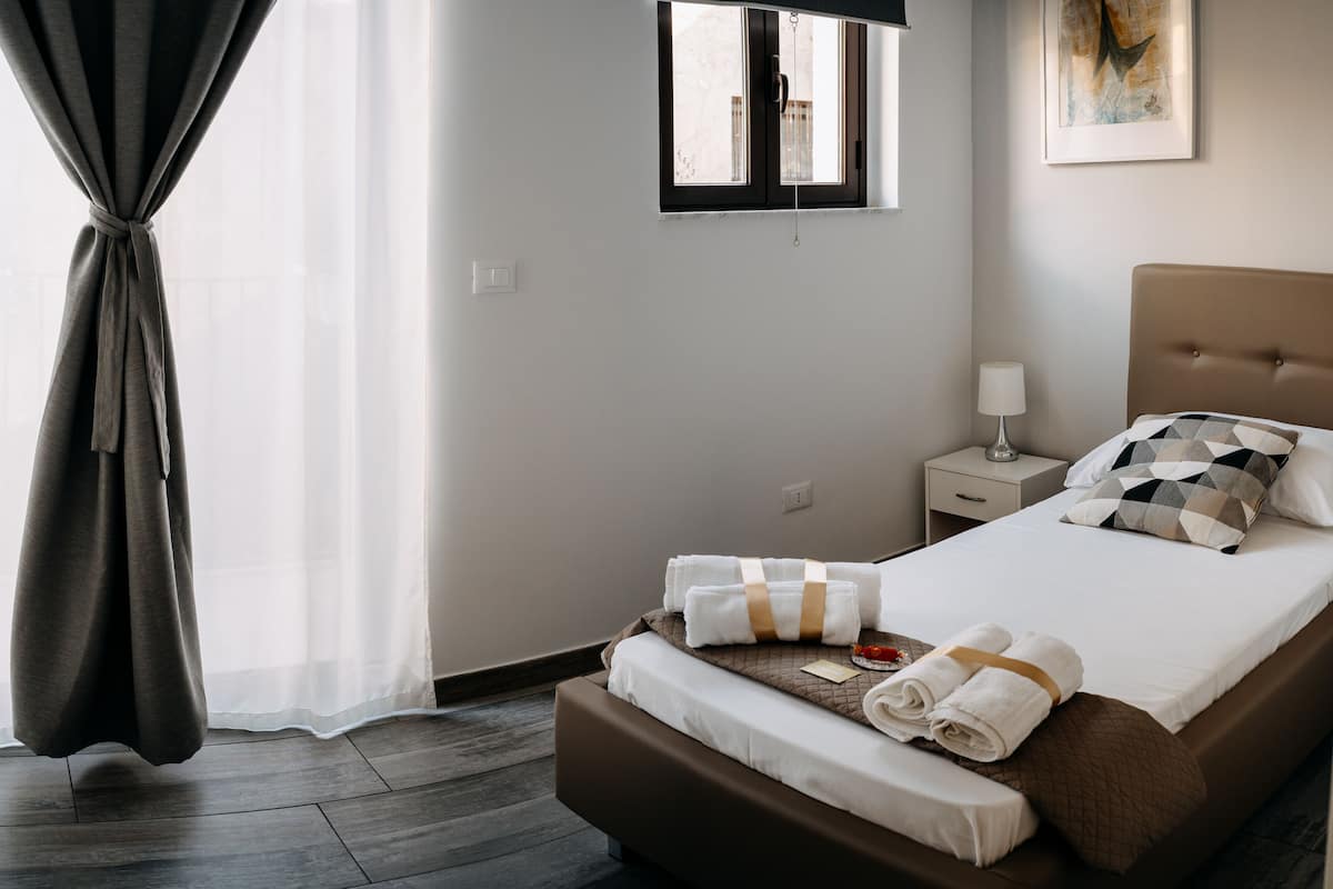 Quarto de hotel com cama, mesa de cabeceira com abajur, janela com vista e cortina.