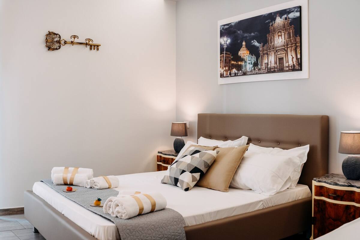 Quarto de hotel moderno com uma cama grande, mesas de cabeceira, um criado-mudo, uma lâmpada e um quadro emoldurado na parede.