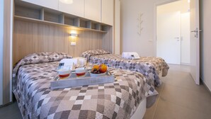 Quarto de hotel com duas camas, cada uma com colcha xadrez, uma bandeja com toalhas e fruta, e prateleiras embutidas acima das camas.