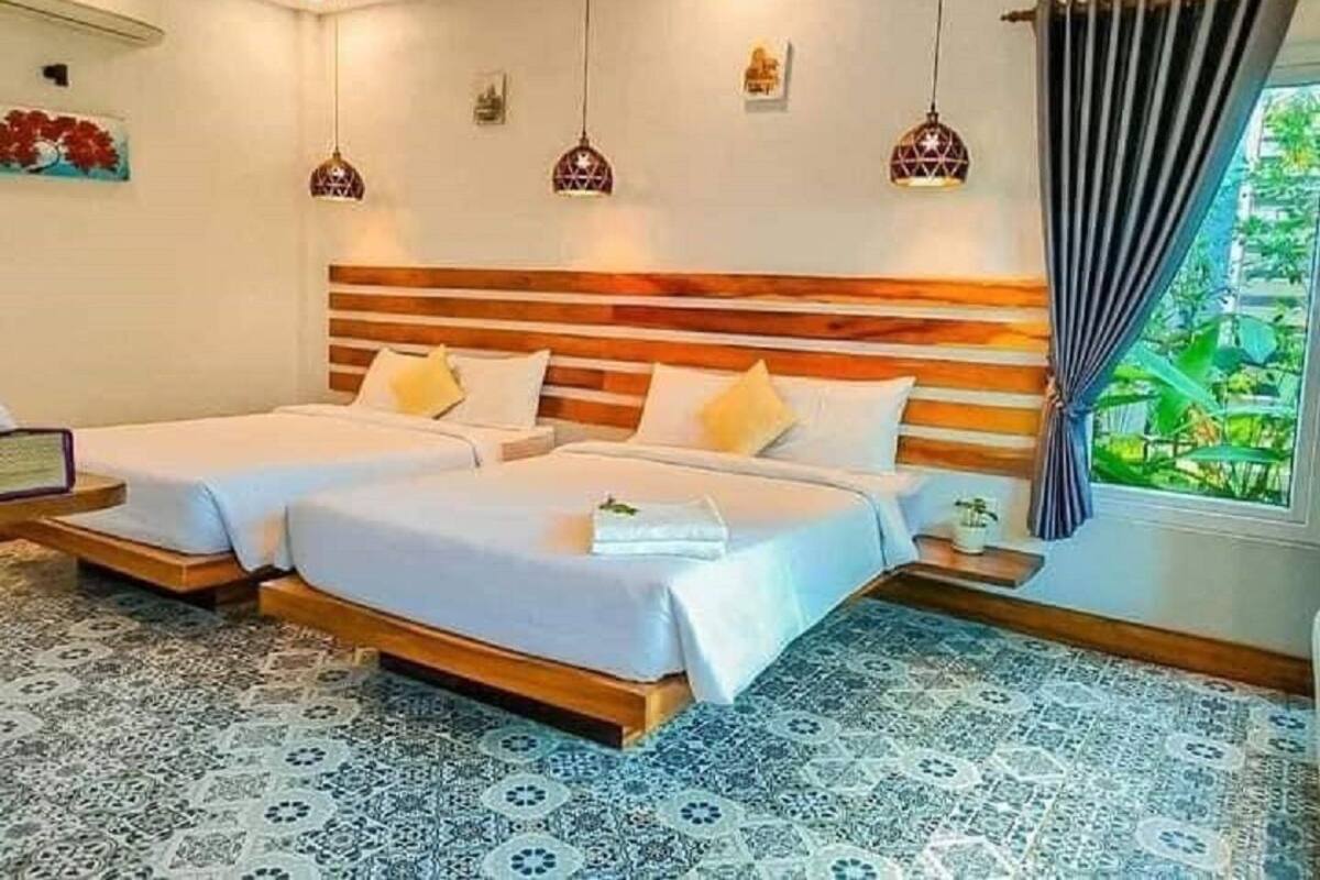 Quarto de hotel com duas camas, cabeceira de madeira, ladrilhos estampados, luminárias pendentes e uma janela com cortinas.
