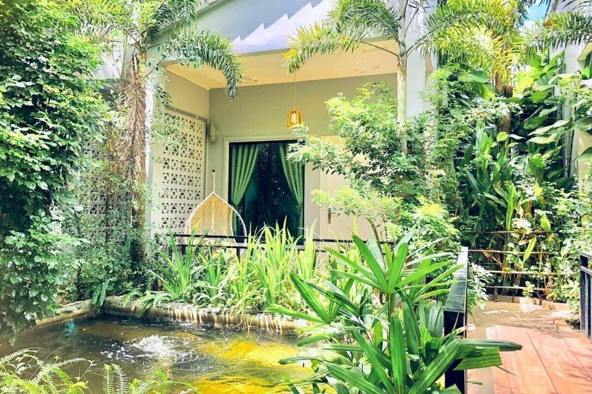 Um jardim exuberante com um lago, um deck de madeira e uma janela com cortinas verdes.