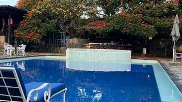 Uma piscina com uma espreguiçadeira, uma árvore com flores vermelhas e um pátio de pedra.