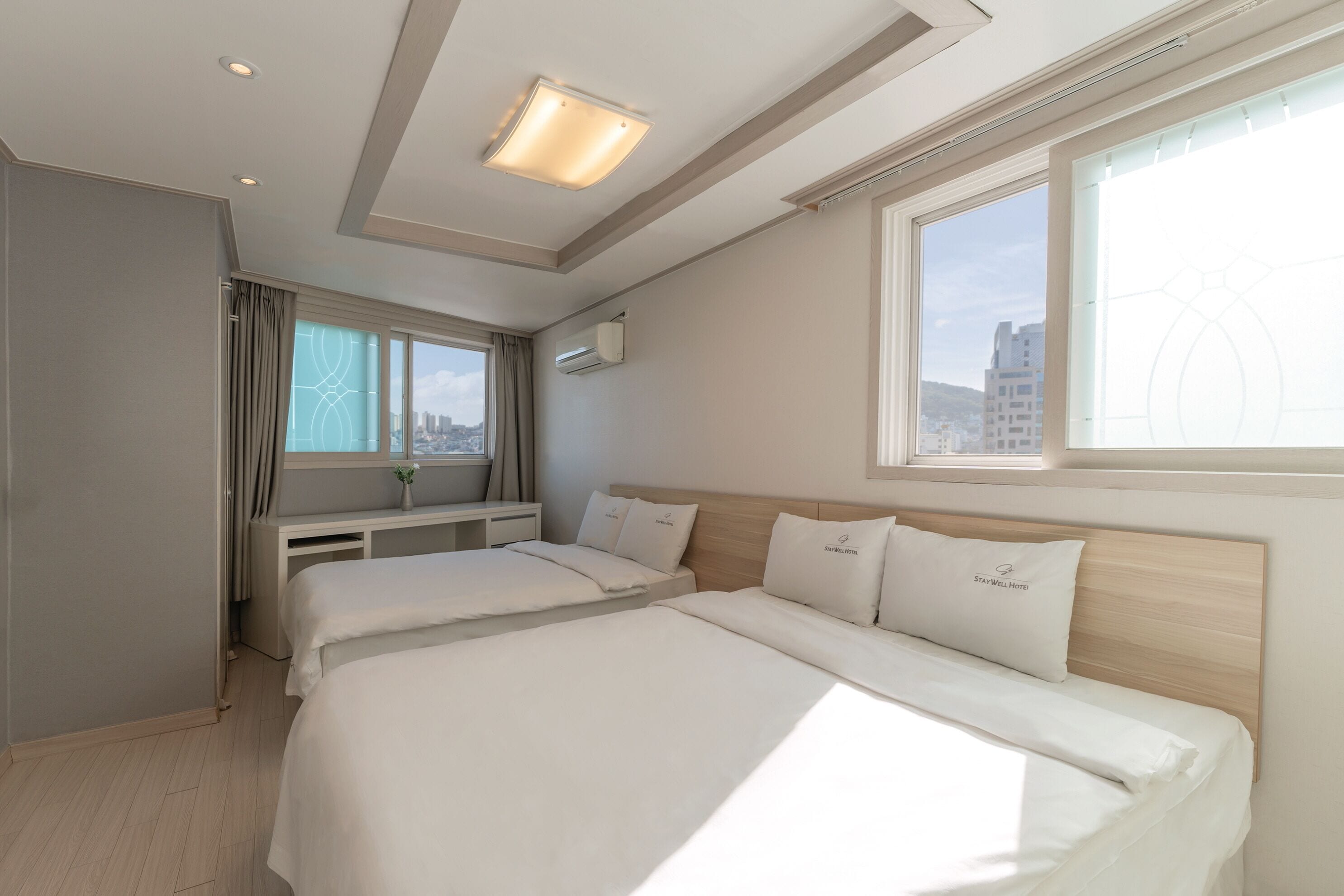 Quarto de hotel com duas camas, janela com vista para a cidade, luminária no teto e ar-condicionado fixado na parede.