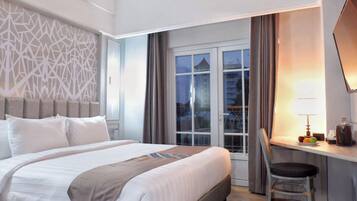 Quarto de hotel com cama, escrivaninha, cadeira e janela com vista para edifícios.