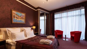 Quarto de hotel com uma cama grande, duas poltronas vermelhas, uma mesa de vidro e um quadro na parede.