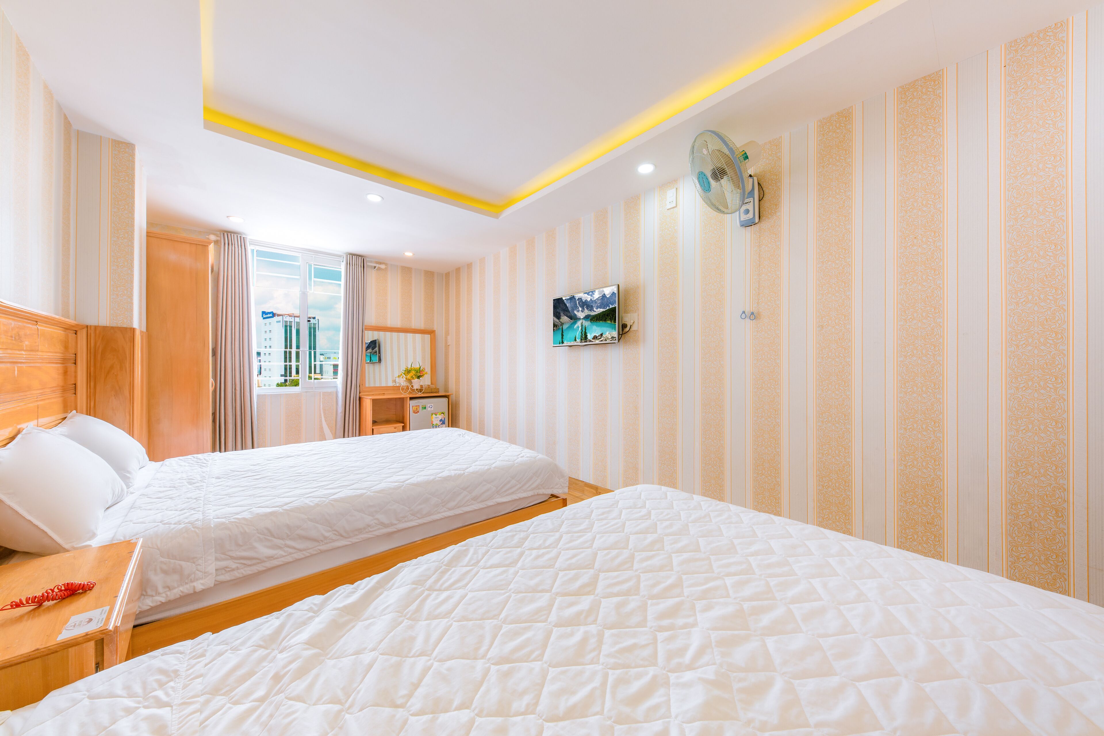 Quarto de hotel com duas camas, uma cabeceira de madeira, mesa de cabeceira, televisão e janela com cortinas.