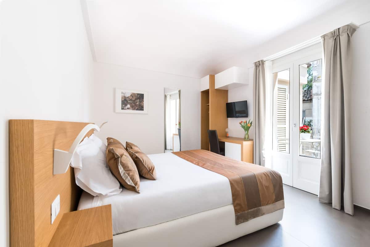 Quarto de hotel moderno com uma cama grande, uma escrivaninha, uma televisão e uma varanda com cortinas.