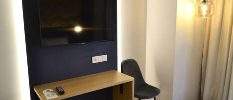 Quarto de hotel com televisão de tela plana na parede, uma escrivaninha de madeira com controle remoto, uma cadeira preta e um lustre.