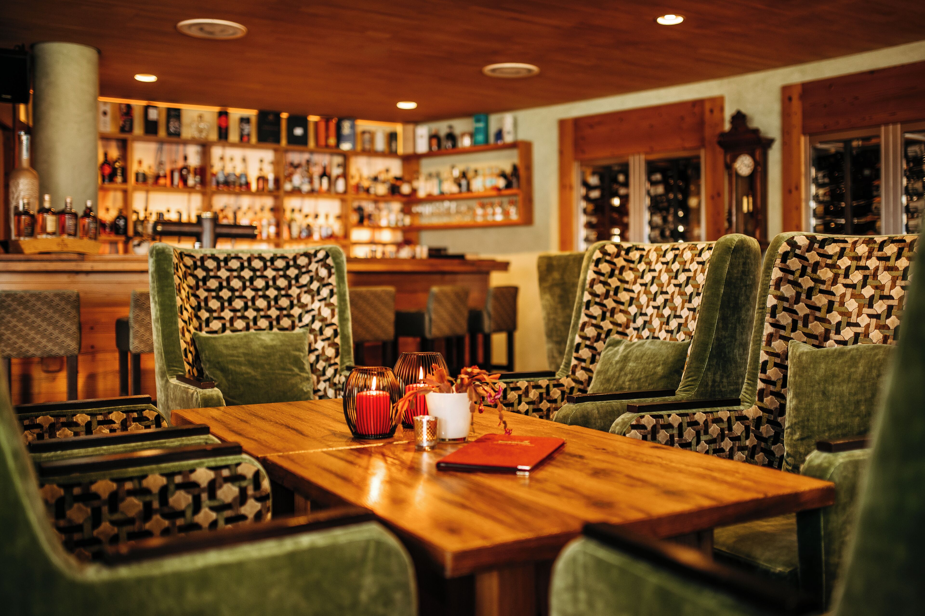 Uma área de bar com uma mesa de madeira, cadeiras estofadas em verde e diversos frascos nas prateleiras.