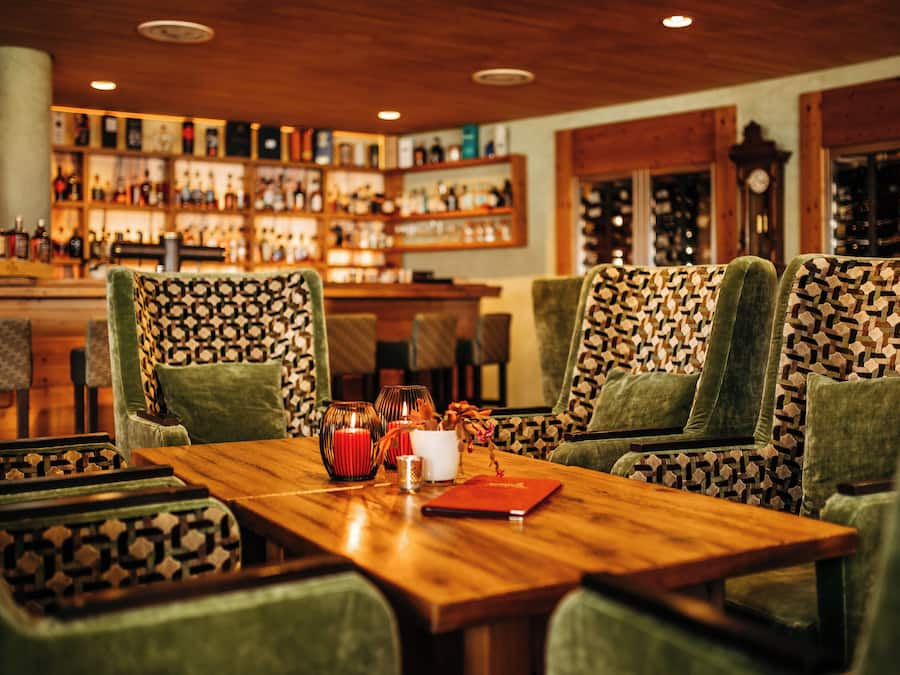 Uma área de bar com uma mesa de madeira, cadeiras estofadas em verde e diversos frascos nas prateleiras.