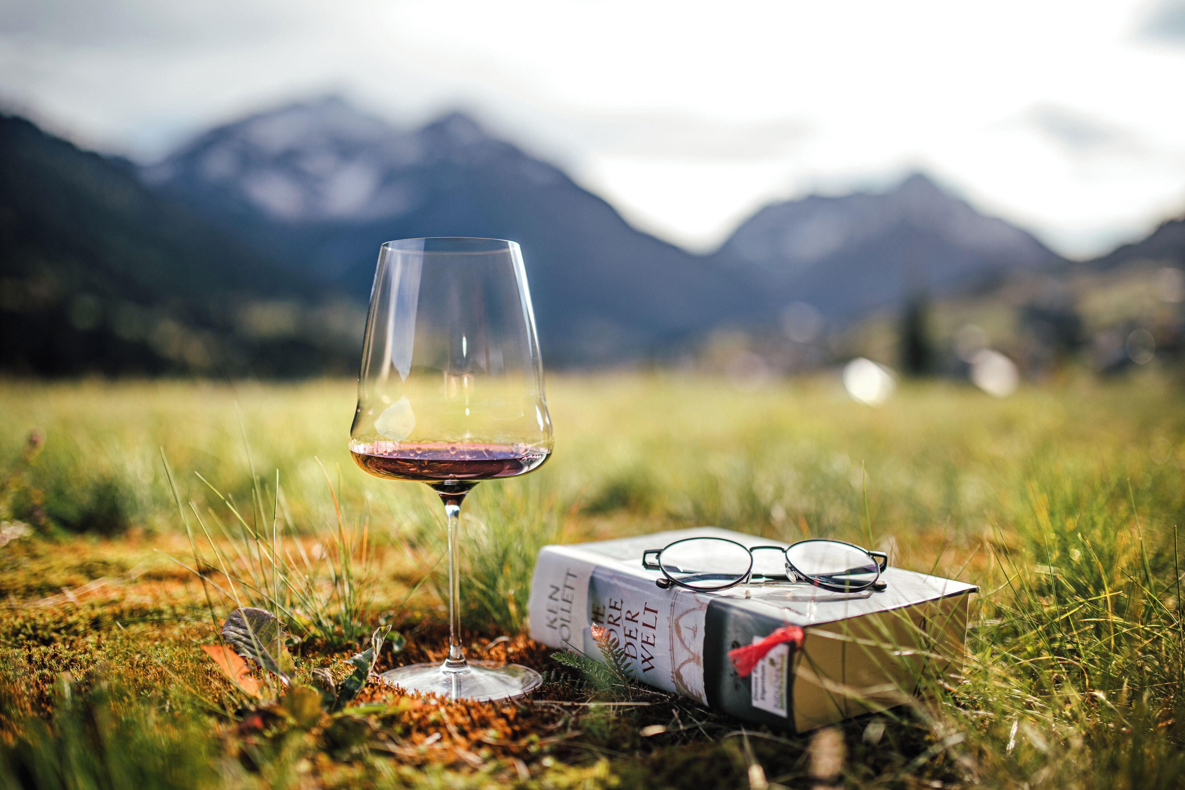 Um copo de vinho tinto, um livro e óculos sobre um campo gramado, com montanhas ao fundo.