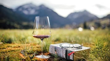 Um copo de vinho tinto, um livro e óculos sobre um campo gramado, com montanhas ao fundo.