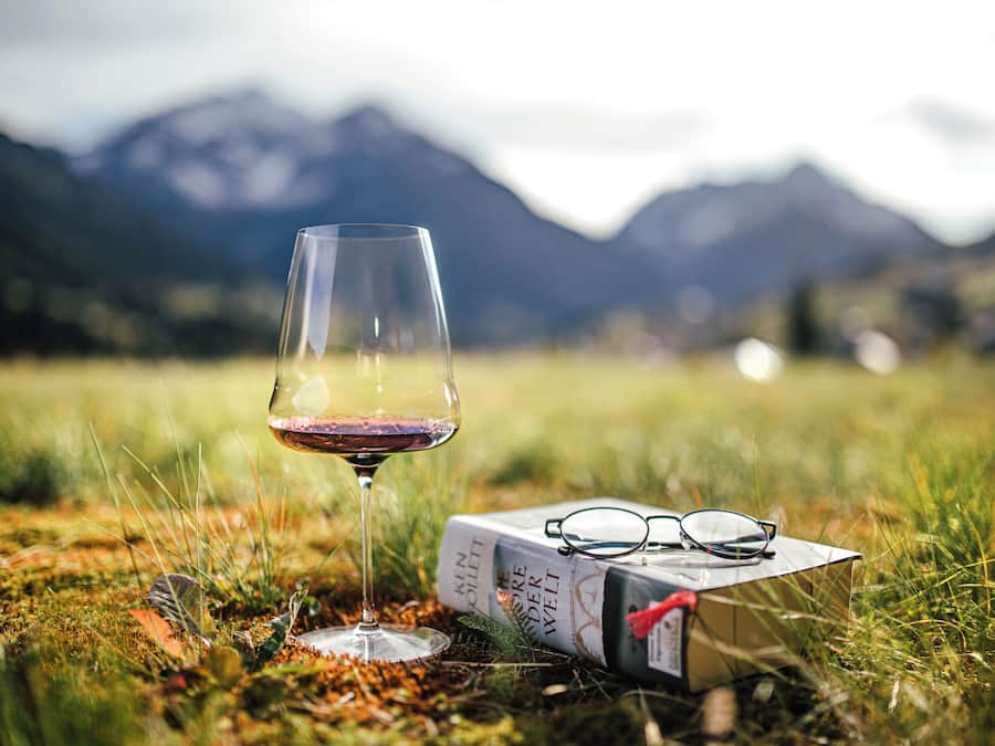 Um copo de vinho tinto, um livro e óculos sobre um campo gramado, com montanhas ao fundo.