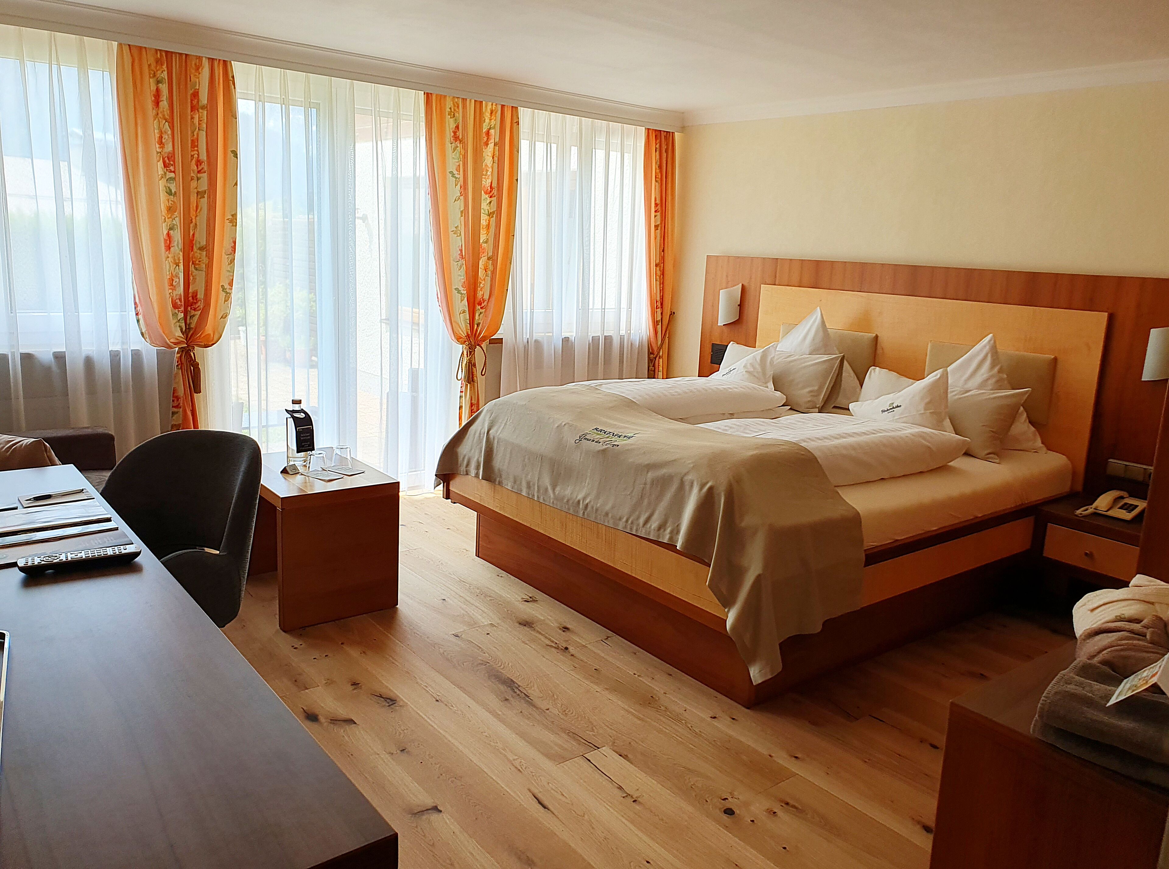 Quarto de hotel com uma cama de madeira, uma escrivaninha com cadeira e uma janela com cortinas.