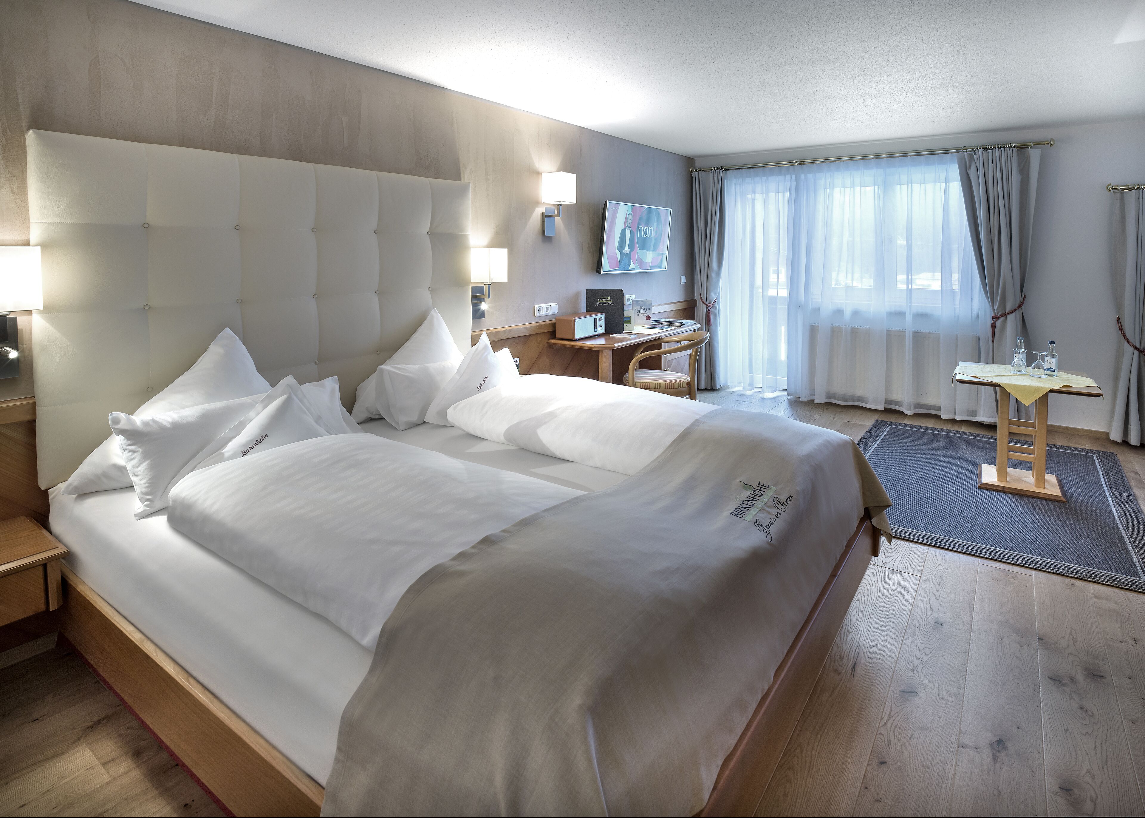 Quarto de hotel com uma cama grande, escrivaninha, cadeira, televisão e janela com cortinas.