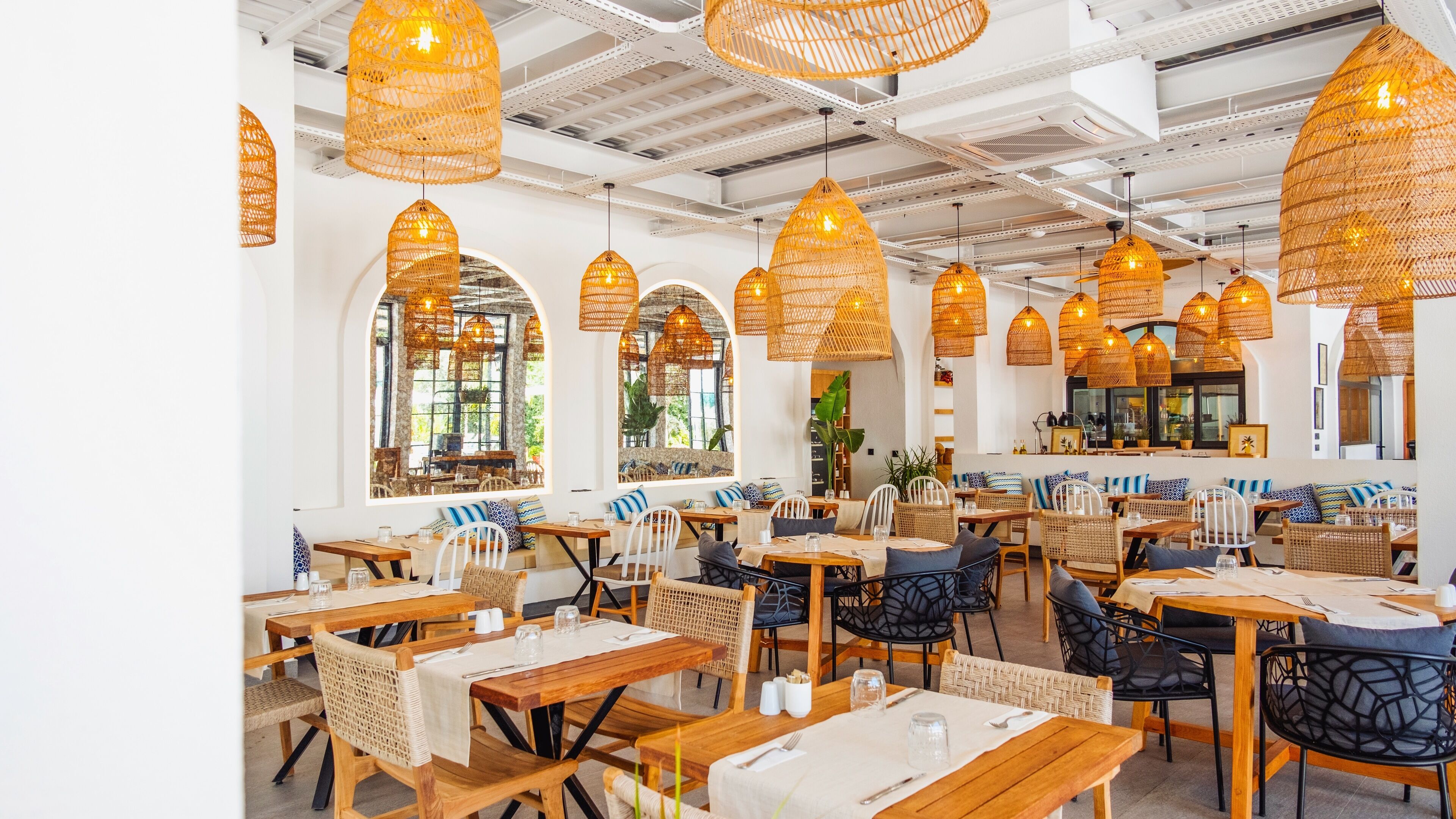 Interior de restaurante com mesas e cadeiras de madeira, luminárias de vime penduradas e janelas amplas.