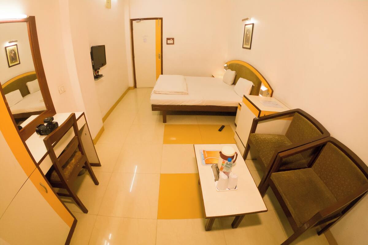 Quarto de hotel compacto com cama, escrivaninha, cadeira e pequena mesa com uma garrafa de água e um controle remoto.