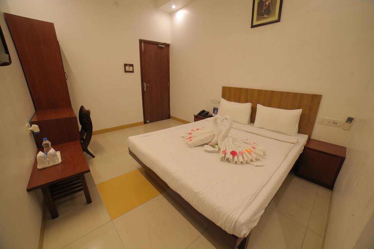 Quarto de hotel com uma cama, mesa de cabeceira de madeira, escrivaninha com produtos de higiene pessoal, uma cadeira e uma porta.