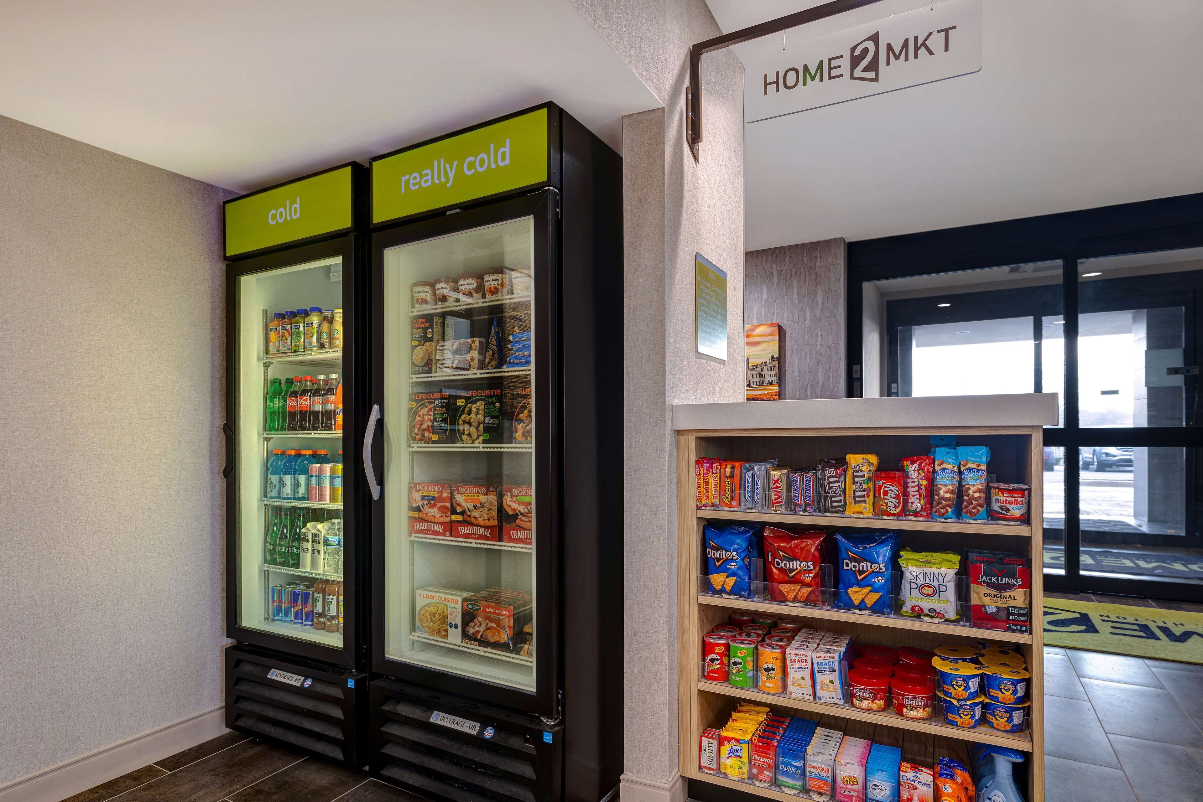 Um saguão de hotel com um refrigerador que exibe bebidas e lanches, com etiquetas indicando 'frio' e 'bem frio'.