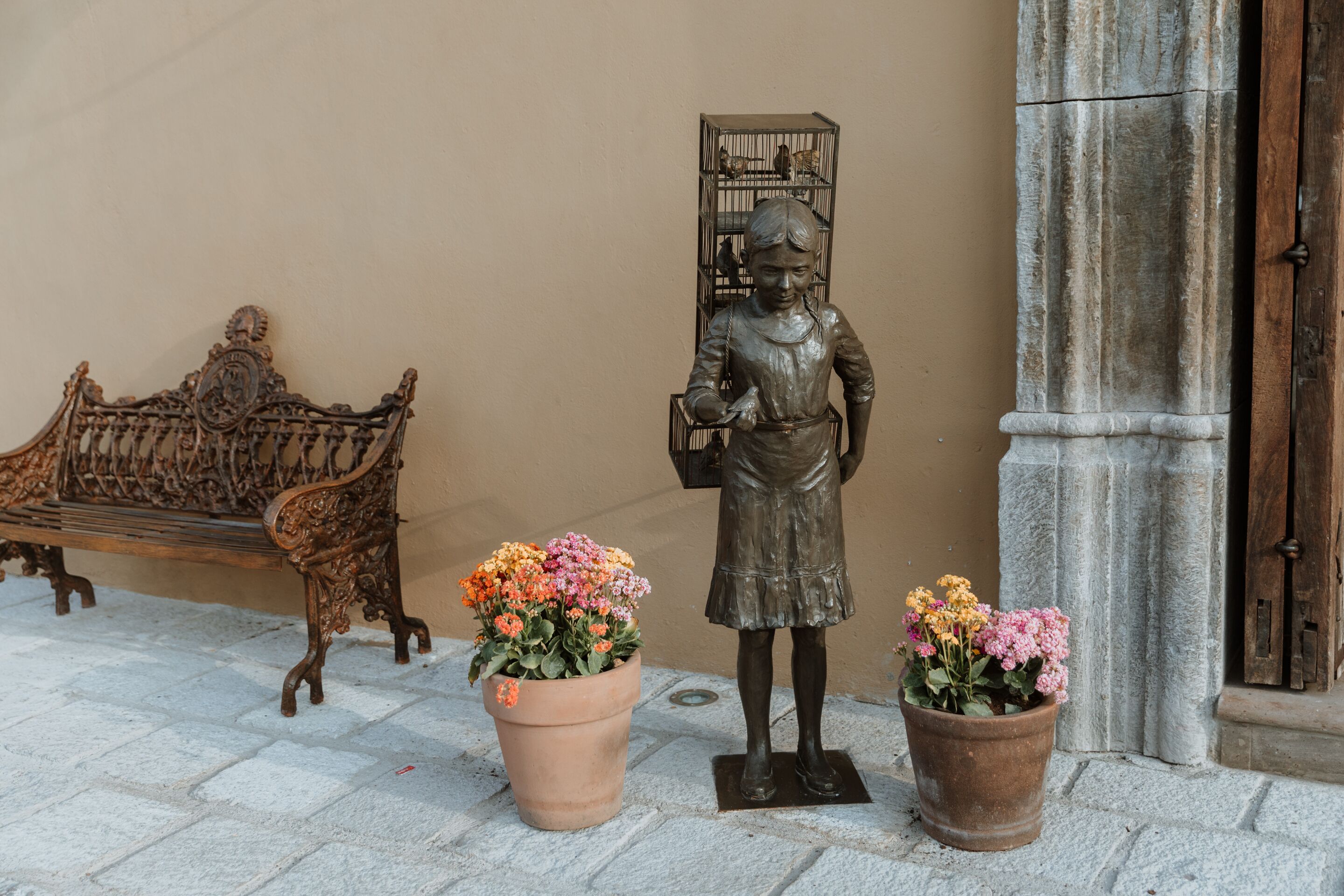 Estátua de mulher segurando uma gaiola de pássaros, com duas plantas em vasos floridos ao lado, em frente a um edifício com um banco e um pilar.