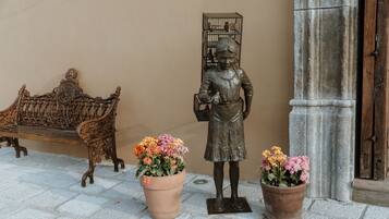 Estátua de mulher segurando uma gaiola de pássaros, com duas plantas em vasos floridos ao lado, em frente a um edifício com um banco e um pilar.