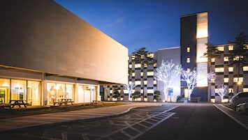 Um edifício moderno com fachada plana e grandes janelas. Há mesas de piquenique do lado de fora e um estacionamento com carros.