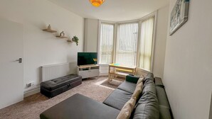Uma sala de estar moderna com uma grande TV de tela plana, um sofá, uma mesa de centro e uma pequena mesa de madeira com uma cadeira.