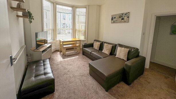 Uma sala de estar com sofá, apoio para os pés e uma mesa. Há uma janela com persianas, um radiador e uma planta suspensa.